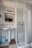 Casa de banho com uma pia branca, espelho, cabine de duche e um caixote de lixo azul.