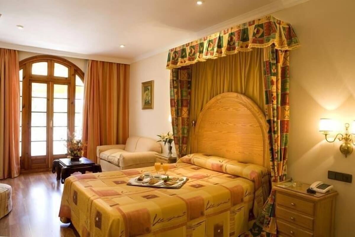 Quarto de hotel com uma cama grande, um espaço de estar com uma cadeira, uma mesinha com um vaso e flores, e uma janela com cortinas.