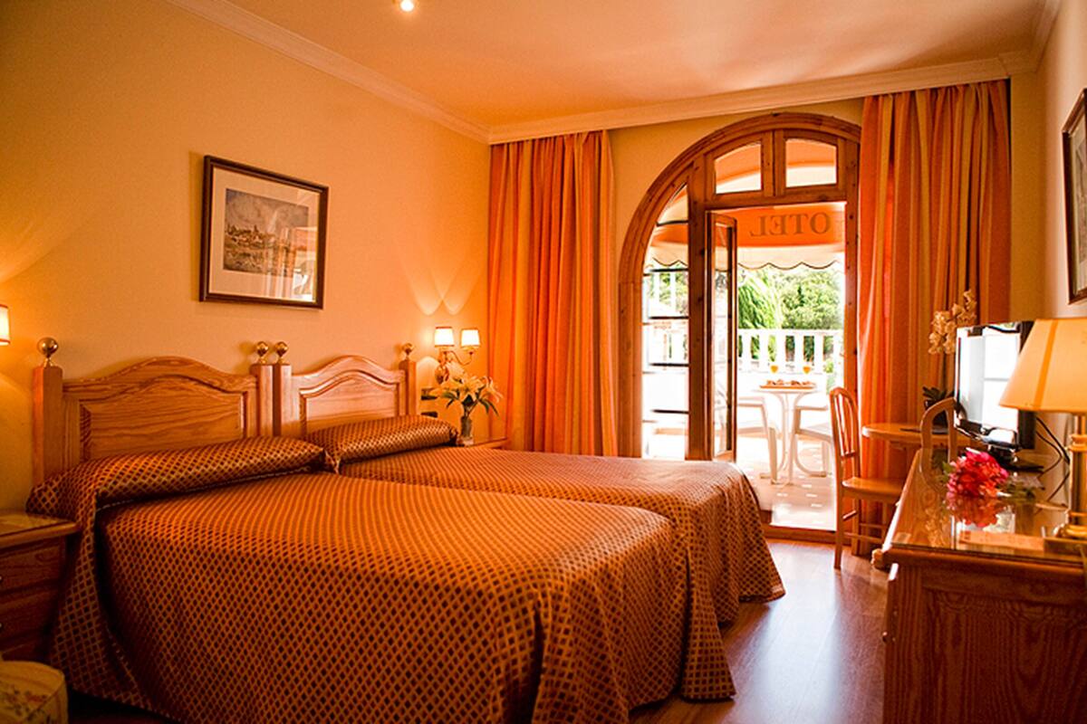 Quarto de hotel com duas camas, cabeceira de madeira, quadro na parede, escrivaninha com abajur e vista para o exterior através de uma porta.