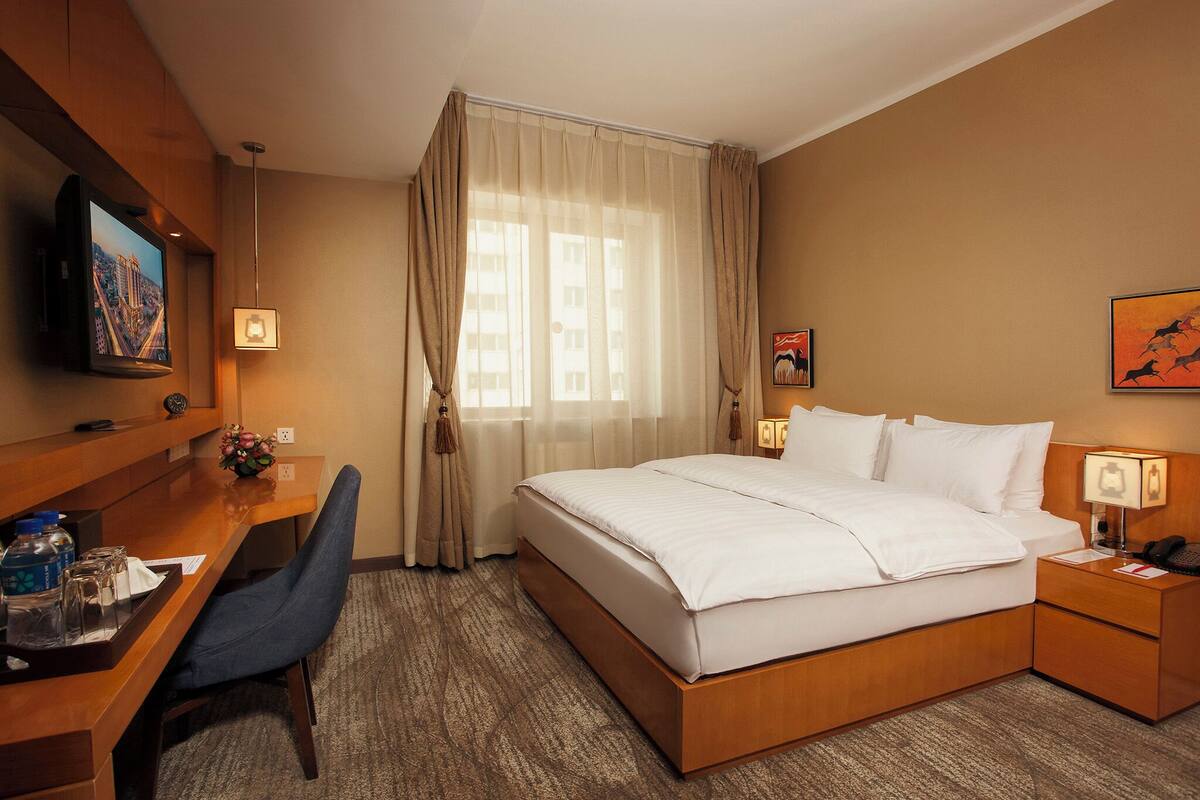 Quarto de hotel com cama, escrivaninha, cadeira, televisão e mesa de cabeceira com luminária.