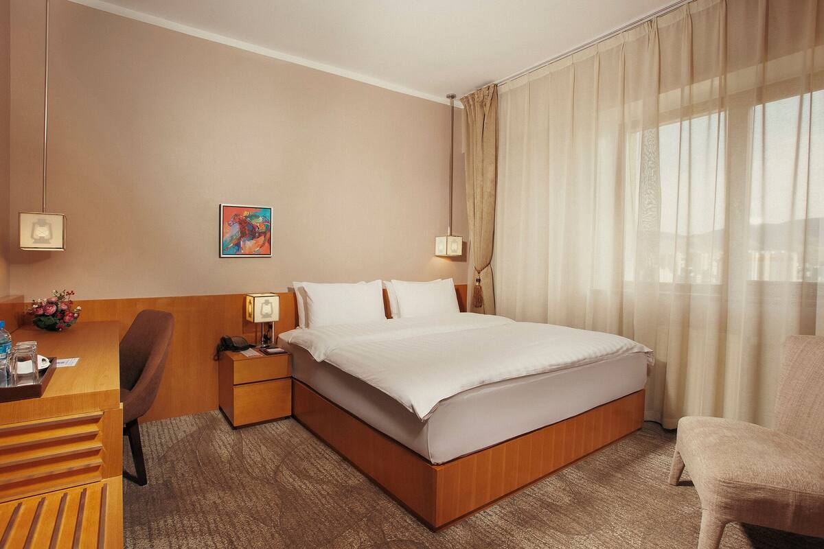 Quarto de hotel com cama grande, escrivaninha com cadeira, mesa de cabeceira, abajur e um quadro na parede.