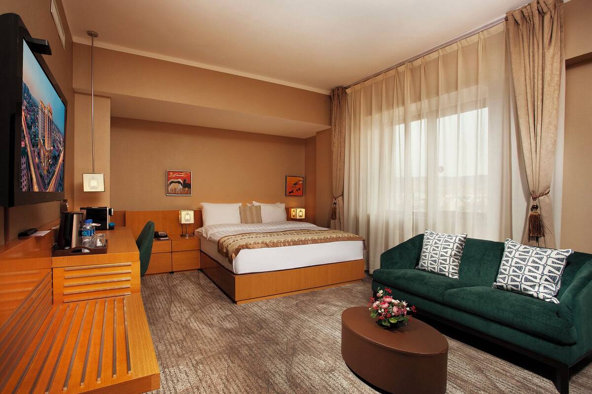Quarto de hotel com uma cama, um sofá verde, uma escrivaninha com computador, televisão e uma janela com cortinas.