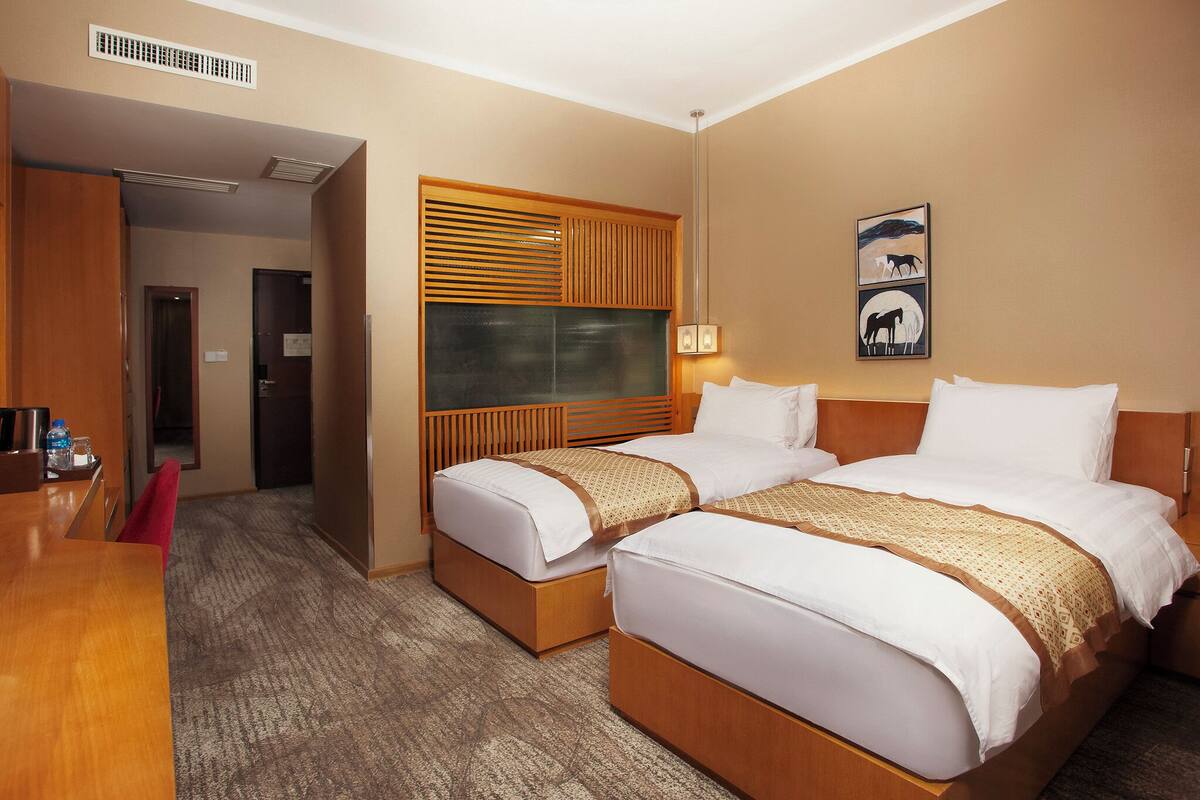 Quarto de hotel com duas camas, cabeceira de madeira, uma janela grande, obra de arte na parede e uma escrivaninha com cadeira.