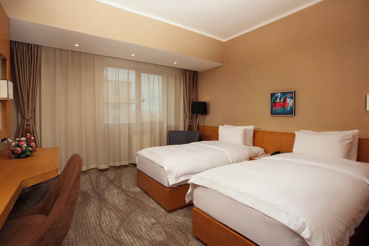 Quarto de hotel com duas camas, uma escrivaninha, uma cadeira, uma televisão e um quadro na parede.
