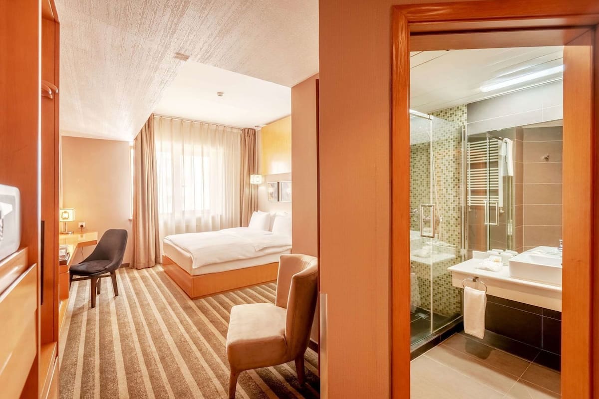 Quarto de hotel com cama, escrivaninha, cadeira, banheiro com chuveiro e espelho.