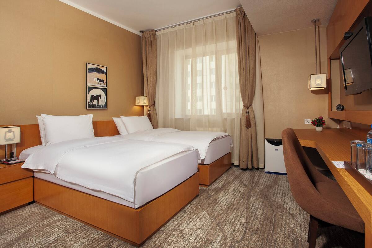 Quarto de hotel com duas camas, uma escrivaninha, uma cadeira, uma televisão e uma janela com cortinas.