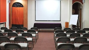 Uma sala de conferências com fileiras de cadeiras, uma tela de projetor e um quadro branco.