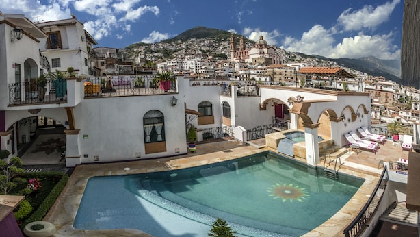 Uma área de piscina com design em formato de sol, rodeada por edifícios brancos com varandas e vista para uma vila na encosta.