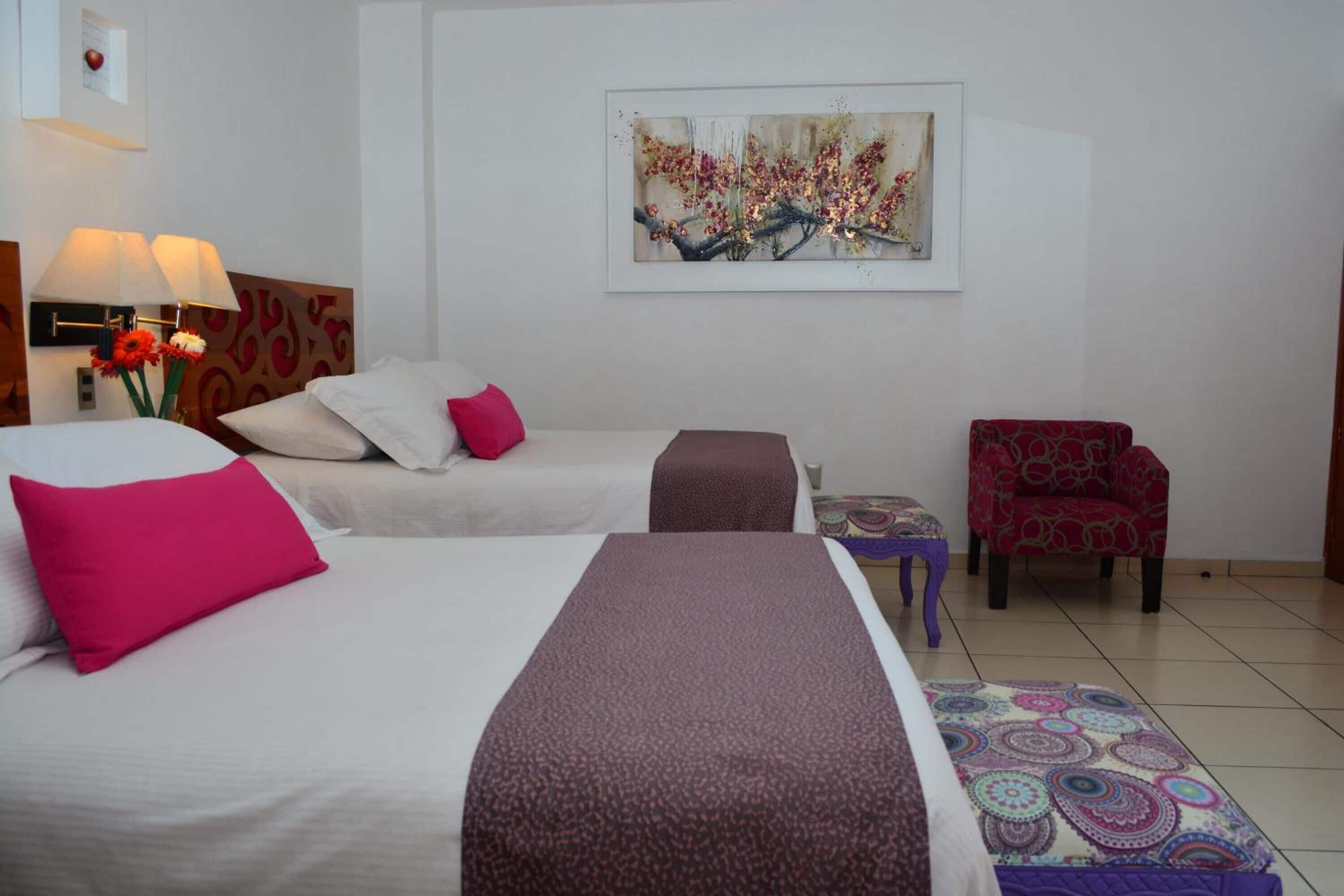 Quarto de hotel com duas camas, uma poltrona vermelha, uma mesinha e um quadro na parede.