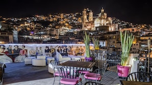 Uma área de estar ao ar livre com mesas e cadeiras, um mural e vista para um horizonte urbano com edifícios iluminados e uma igreja imponente.