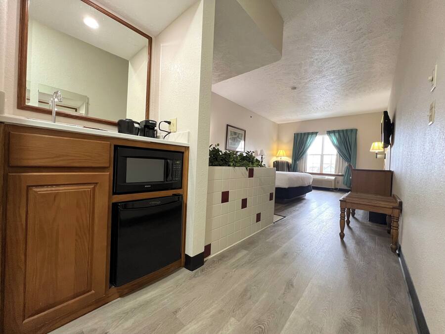 Quarto de hotel com cozinha compacta, cama, mesa de cabeceira, cadeira e janela com cortinas.