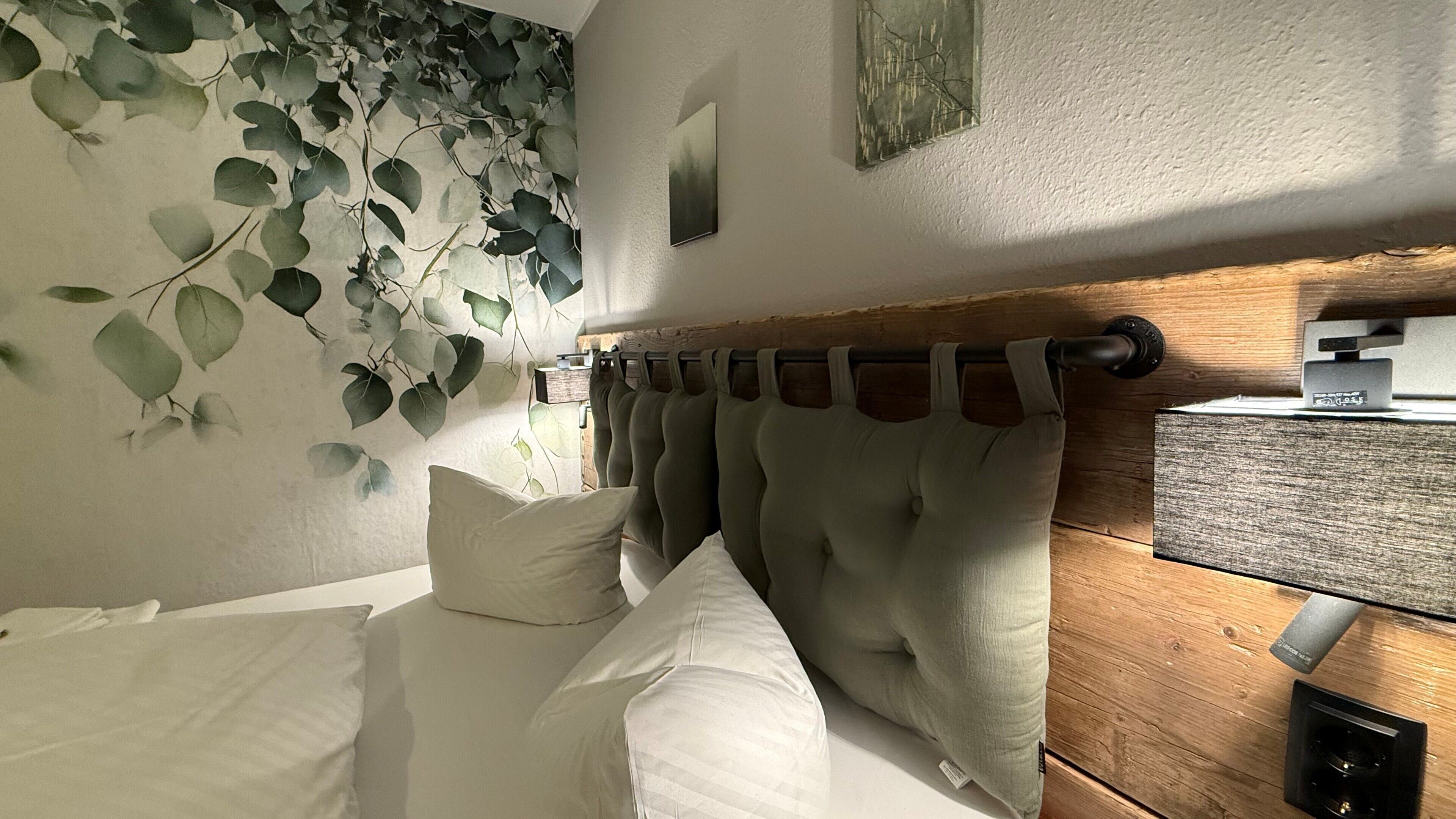 Uma cama bem arrumada com lençóis brancos, cabeceira com painel de madeira e parede decorada com um padrão de folhas.
