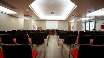 Uma sala de conferências com fileiras de cadeiras, uma tela de projetor e um amplo claraboia.