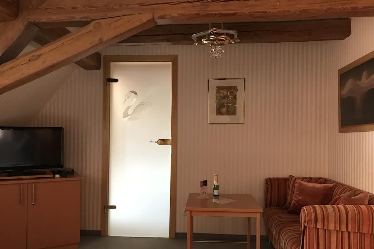 Quarto com televisão sobre um armário de madeira, uma mesinha com uma garrafa, um sofá e uma porta com interruptor de luz.