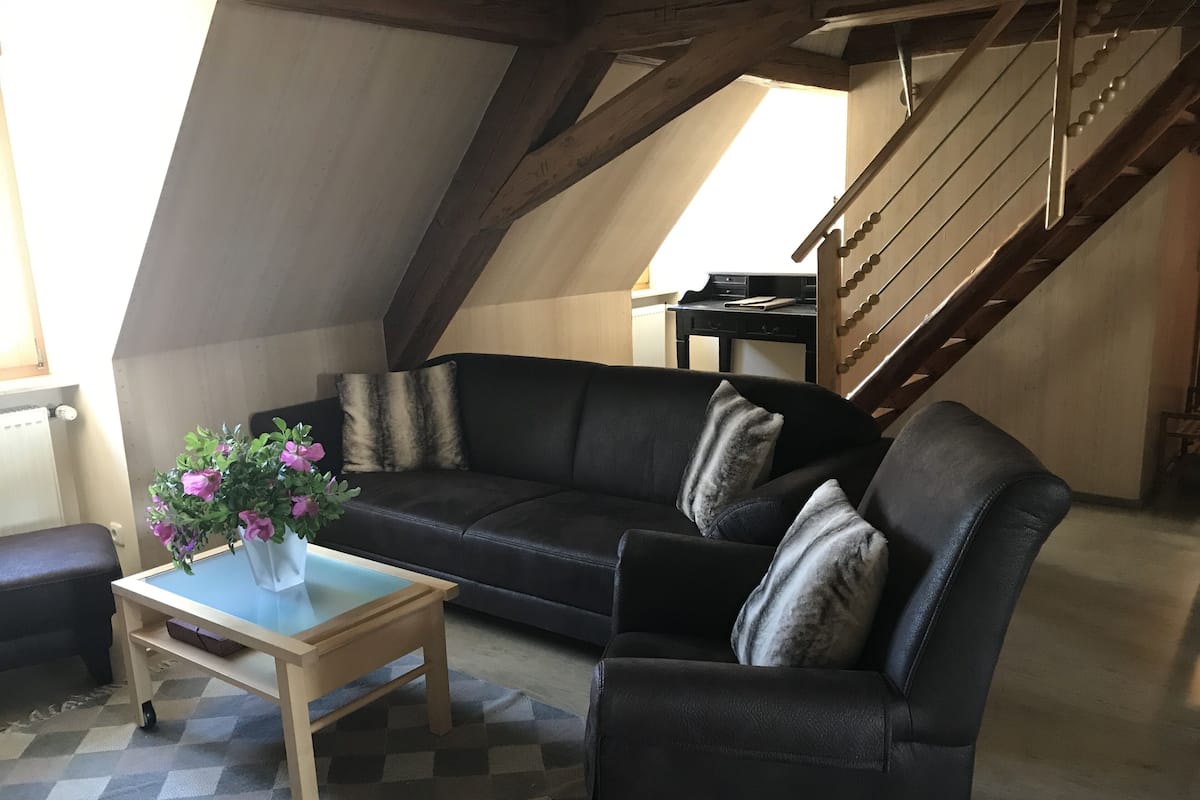 Uma sala de estar moderna com um sofá de couro preto, uma mesa de centro de madeira com um vaso de flores e um tapete xadrez. Uma escada com corrimão de madeira é visível ao fundo.