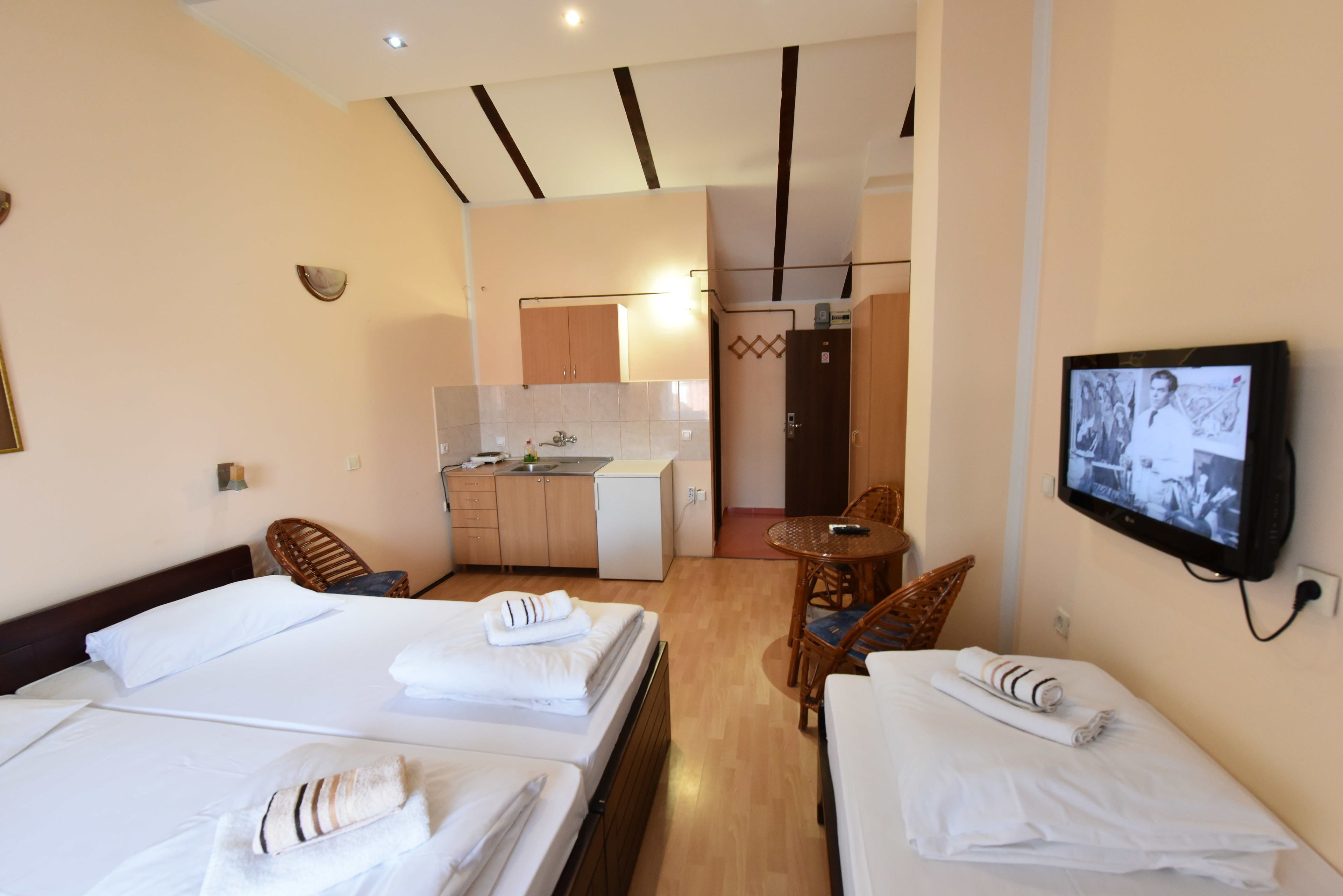 Quarto de hotel com duas camas, cozinha compacta, área de refeições e televisão na parede.