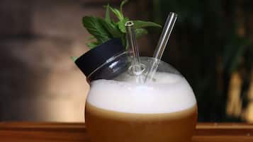 Um copo de vidro transparente com tampa preta e canudo, cheio de uma bebida espumosa, apoiado em uma superfície de madeira com uma planta verde desfocada ao fundo.