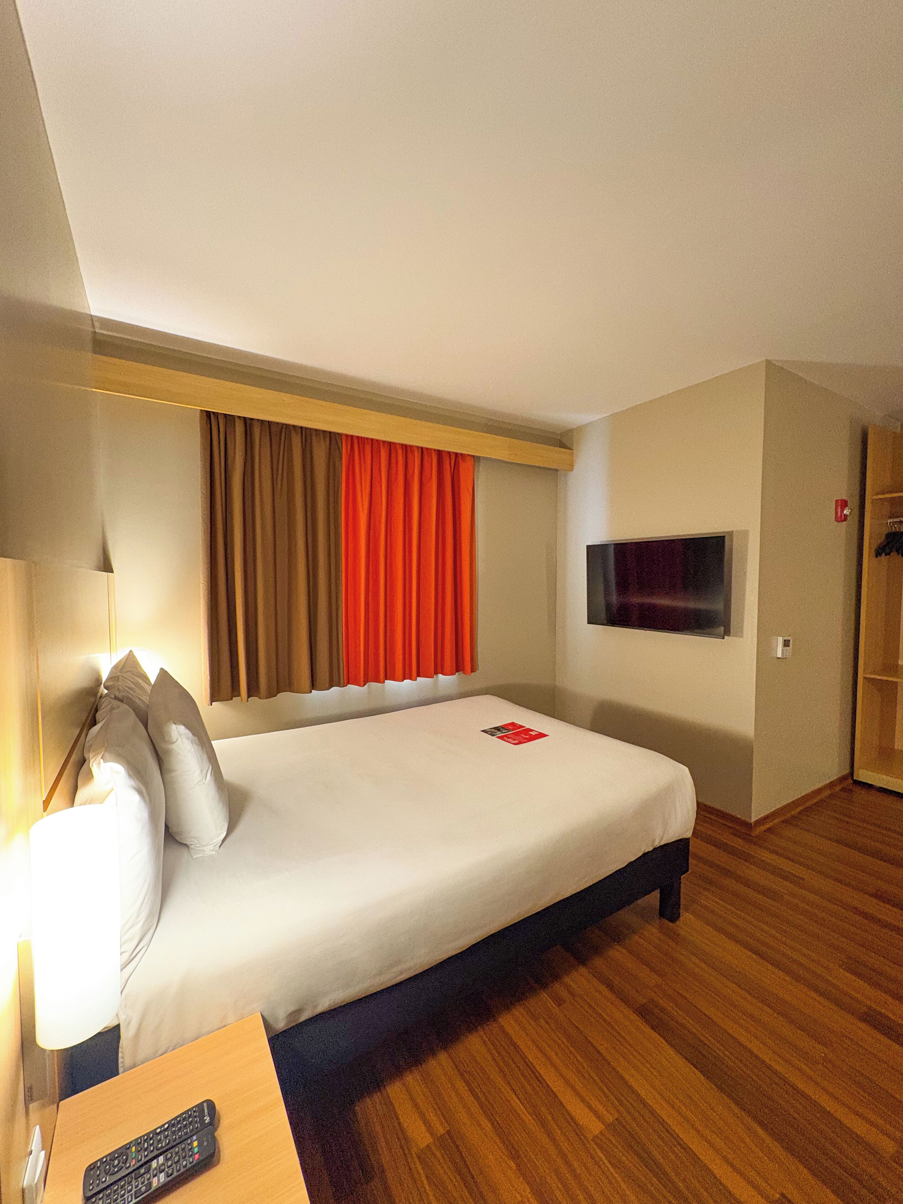 Quarto de hotel com cama, televisão fixada na parede, piso de madeira e janela com cortinas.