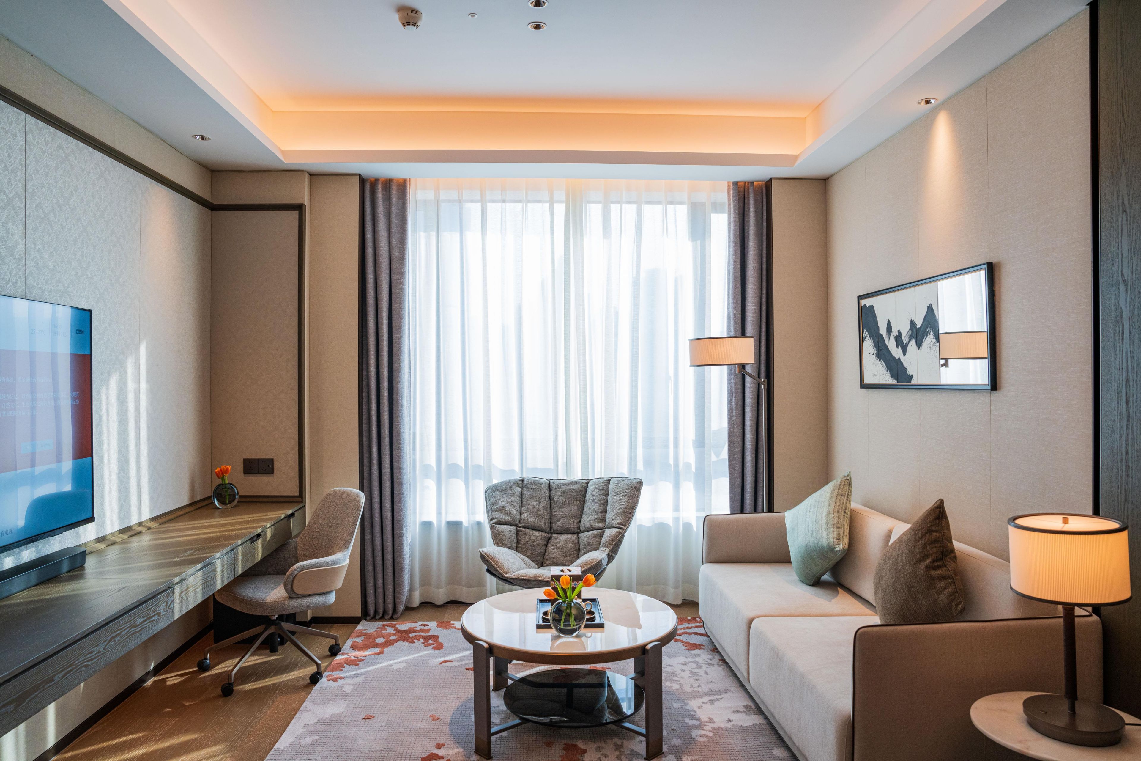 Quarto de hotel moderno com televisão de tela plana, sofá, poltrona, mesa de centro e luminária.