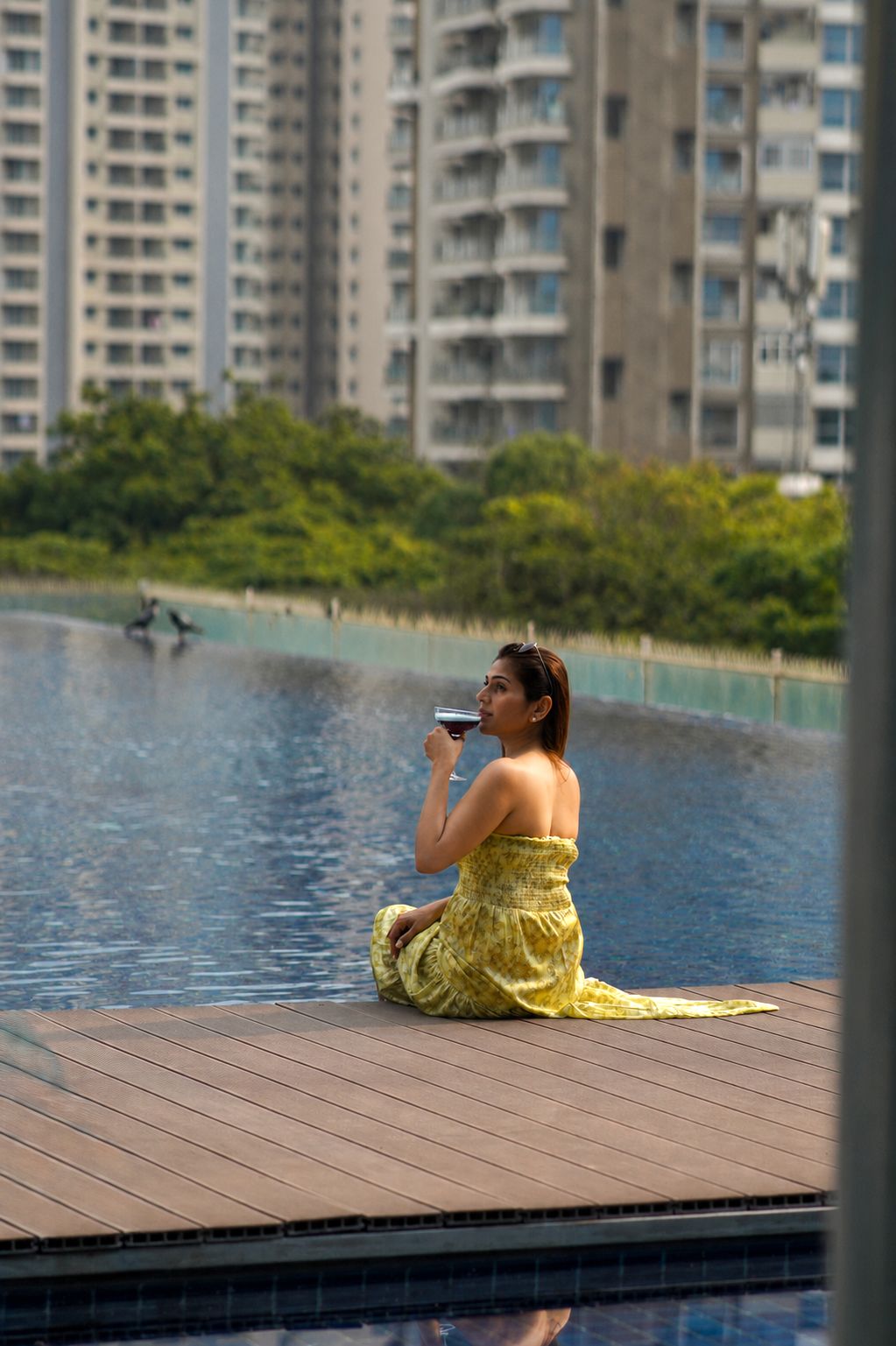 Uma pessoa sentada à beira da piscina, com vista para edifícios altos ao fundo.
