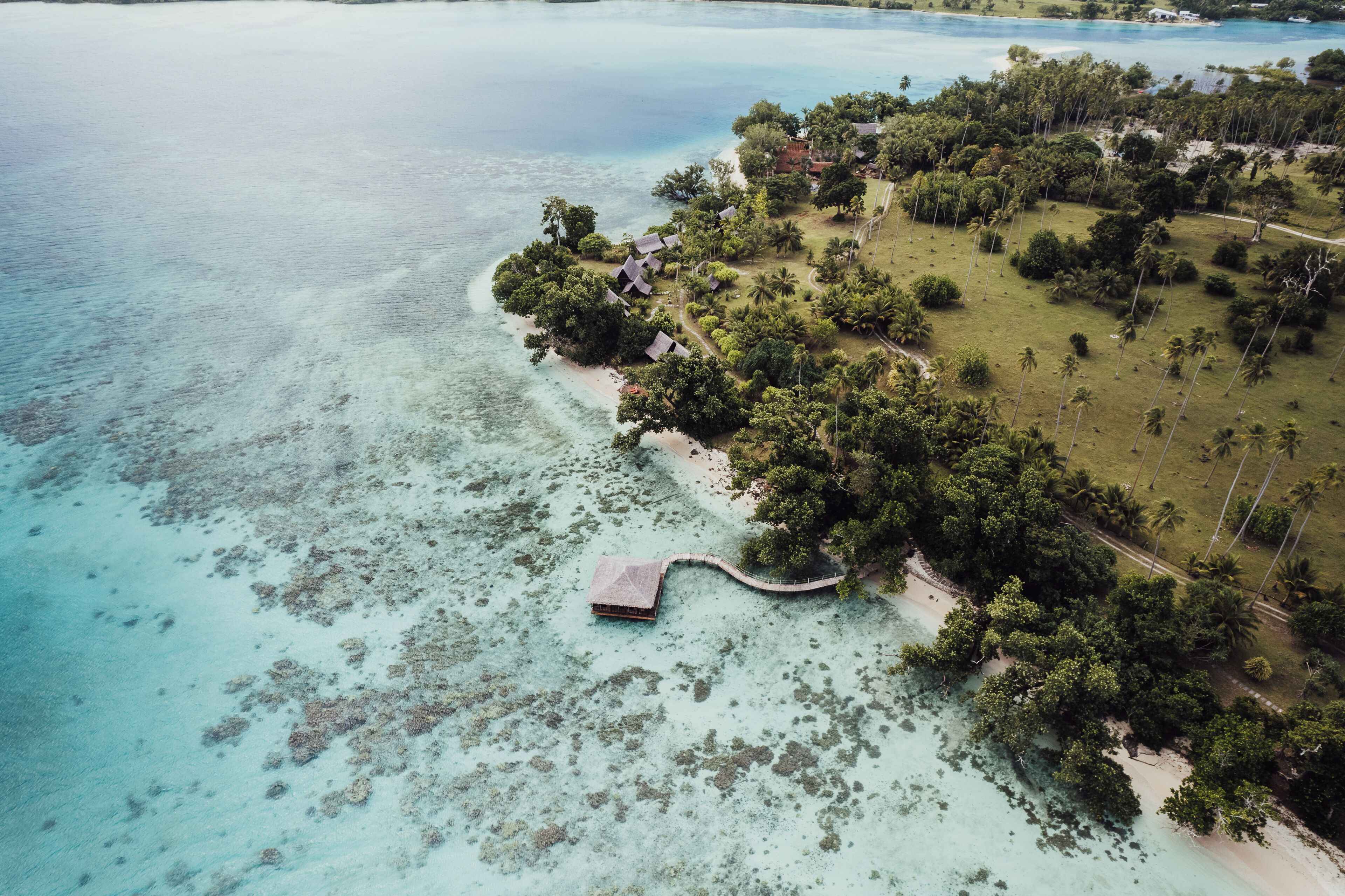 Vista aérea de uma ilha tropical com águas turquesa cristalinas, uma pequena praia e algumas construções cercadas por vegetação exuberante.