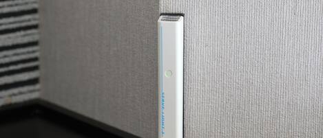 Um dispositivo com indicador luminoso azul, possivelmente uma estação de carregamento ou fonte de energia, apoiado em uma superfície escura contra uma parede texturizada.