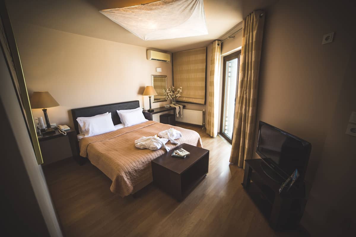 Quarto de hotel com uma cama grande, mesa de cabeceira com luminária, televisão e janela com cortinas.