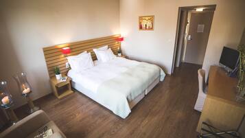 Quarto de hotel com uma cama grande, luminárias laterais, uma escrivaninha com um computador e uma pequena planta.