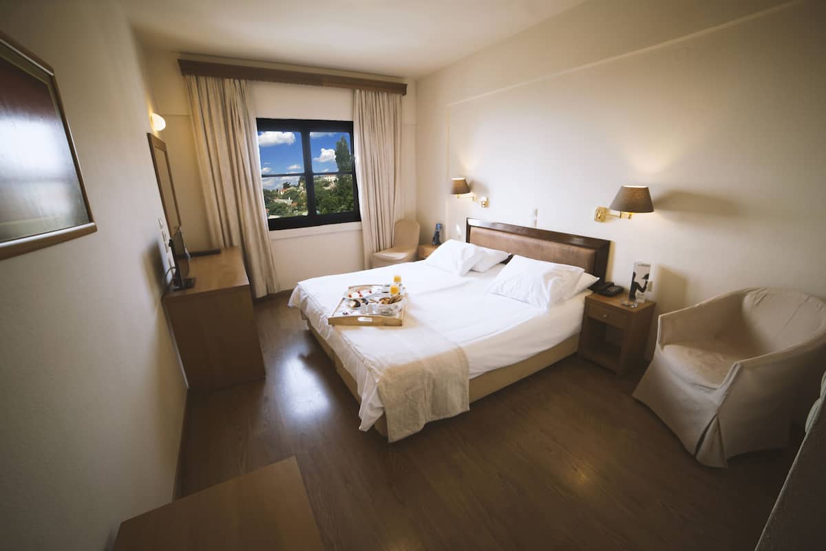 Quarto de hotel com uma cama grande, uma cadeira, mesa de cabeceira, abajur e janela com cortinas.