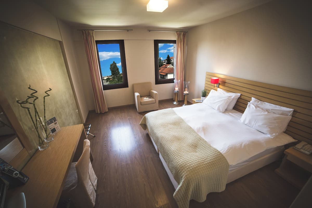 Quarto de hotel com uma cama grande, uma escrivaninha com cadeira, uma mesinha de centro com abajur e duas janelas com cortinas.