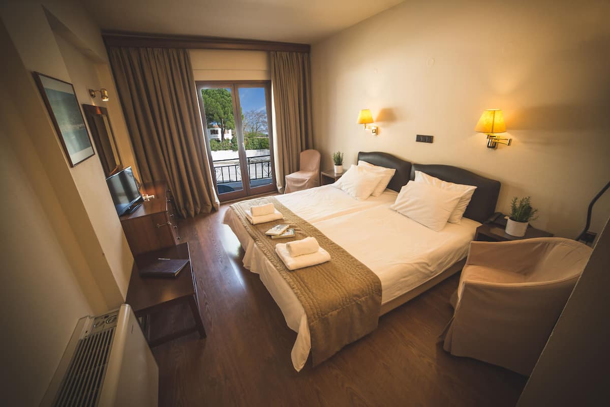 Quarto de hotel com cama, duas luminárias de cabeceira, uma cadeira, uma mesinha, televisão e janela com cortinas.
