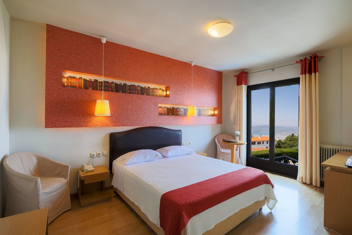Quarto de hotel com uma cama grande, uma cadeira, uma escrivaninha, uma mesa de cabeceira e vista para a cidade através de uma janela ampla.