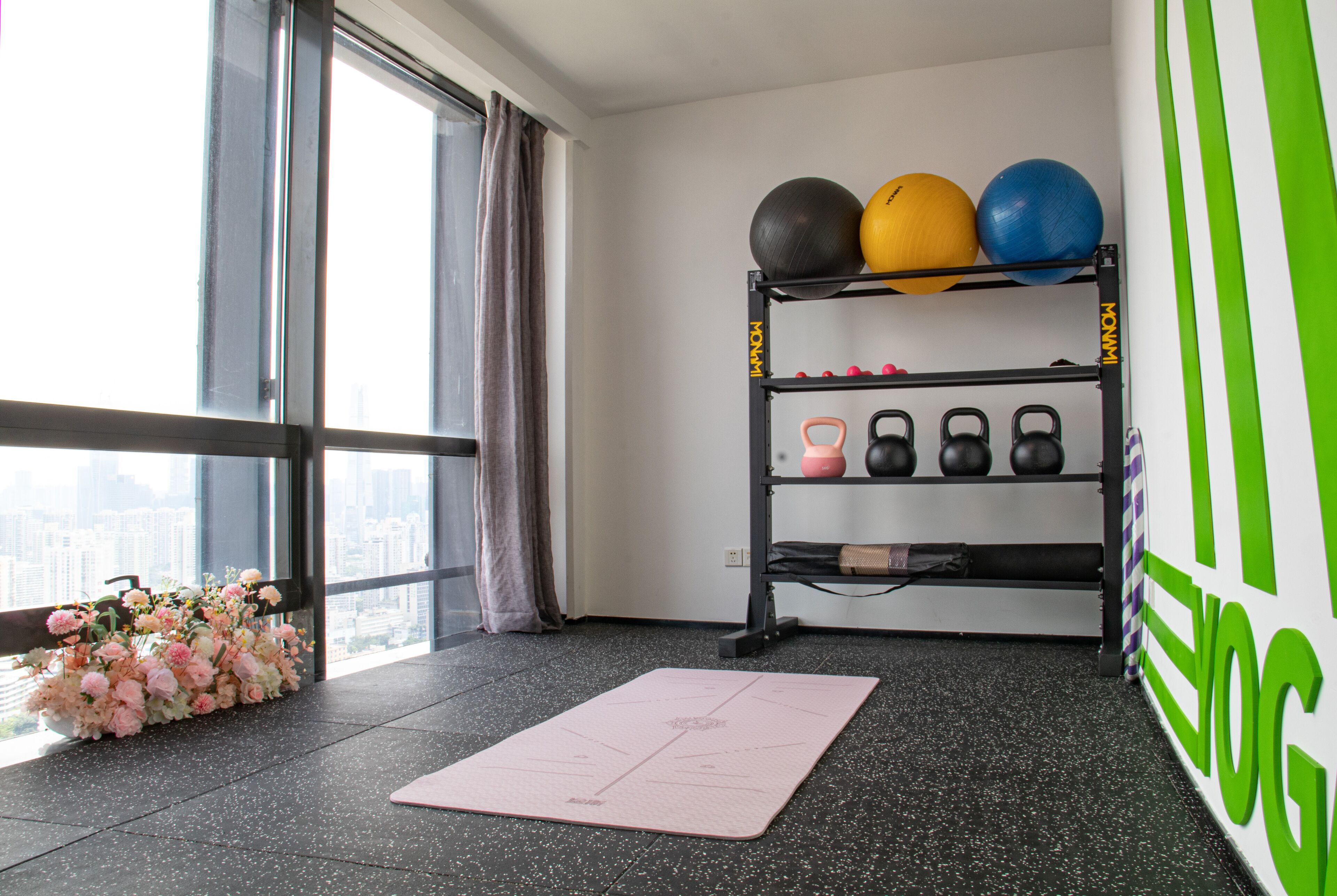 Quarto com uma janela grande, um suporte de exercícios preto com pesos, um tapete de yoga rosa e um arranjo floral.