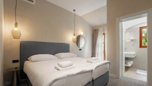 Quarto de hotel moderno com uma cama grande, luminárias laterais, um espelho redondo e vista para o banheiro por uma porta aberta.