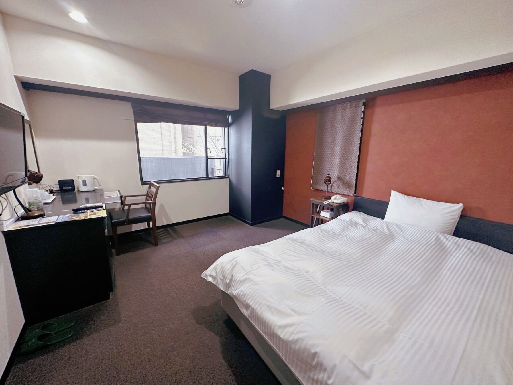 Quarto de hotel com cama, escrivaninha, cadeira e janela.