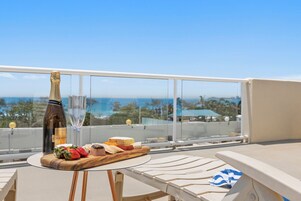 Varanda com mesa posta para refeição, uma garrafa de vinho e um copo. A varanda oferece uma vista panorâmica do oceano e da costa ao longe.