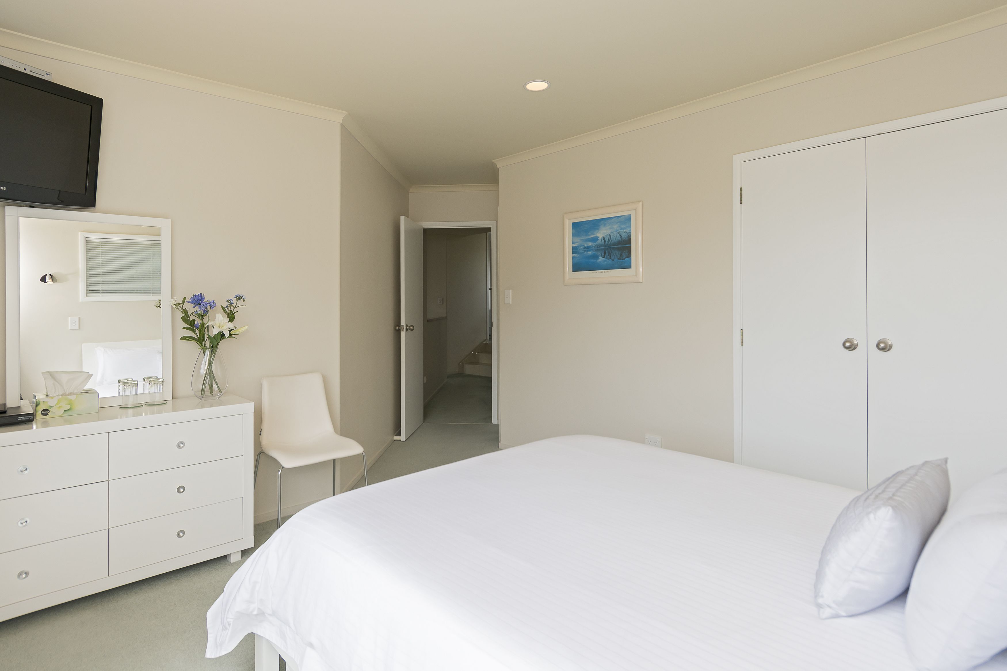 Quarto com cama, cômoda, cadeira, televisão e porta que dá para outro espaço.