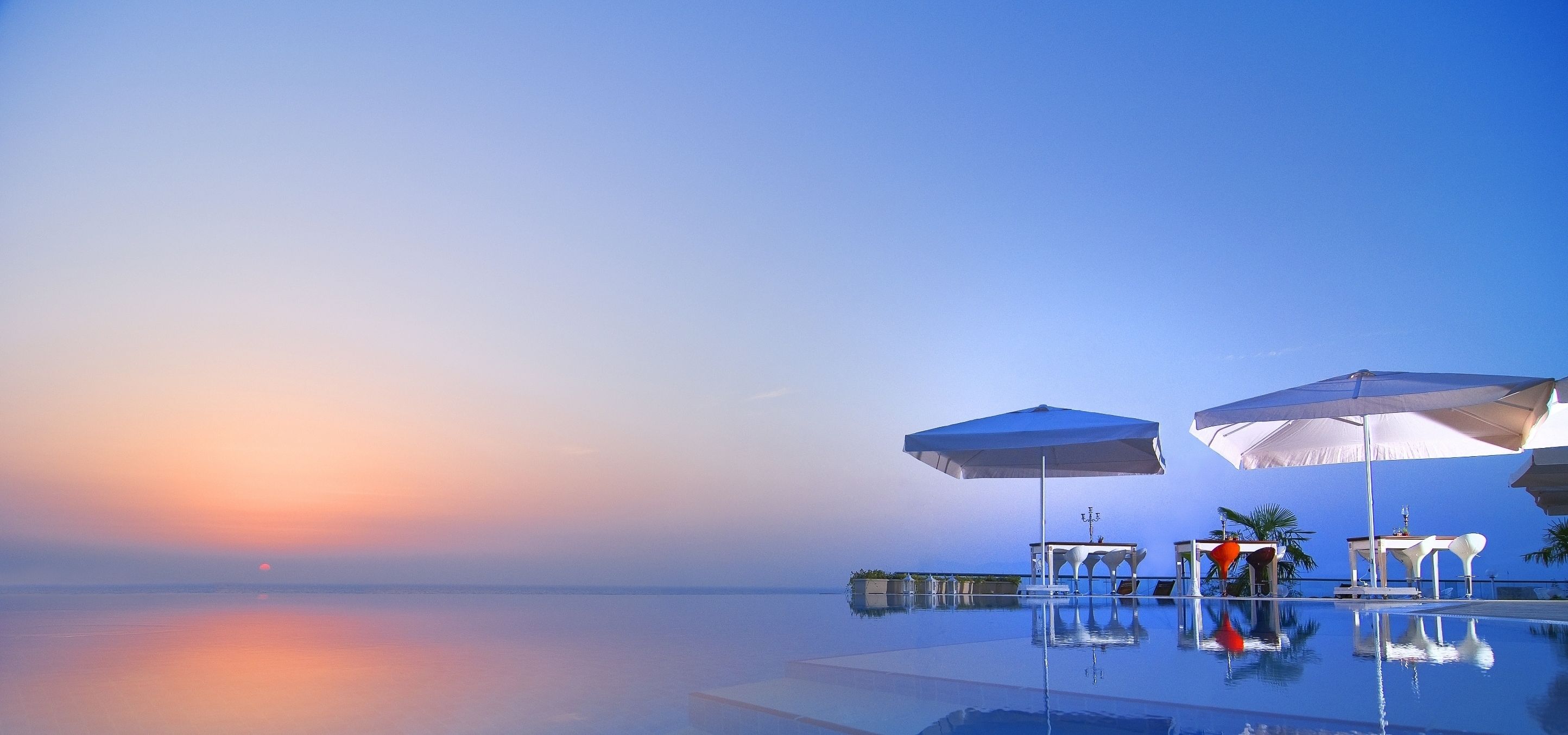 Uma piscina infinita com vista para o pôr do sol, dois guarda-sóis e espreguiçadeiras.