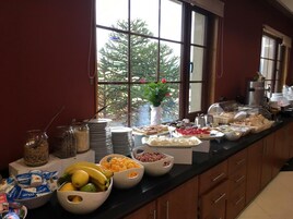 Disposição de buffet com diversas opções de comida, incluindo frutas, sobremesas e bebidas, dispostas em um balcão com vista para o verde lá fora.