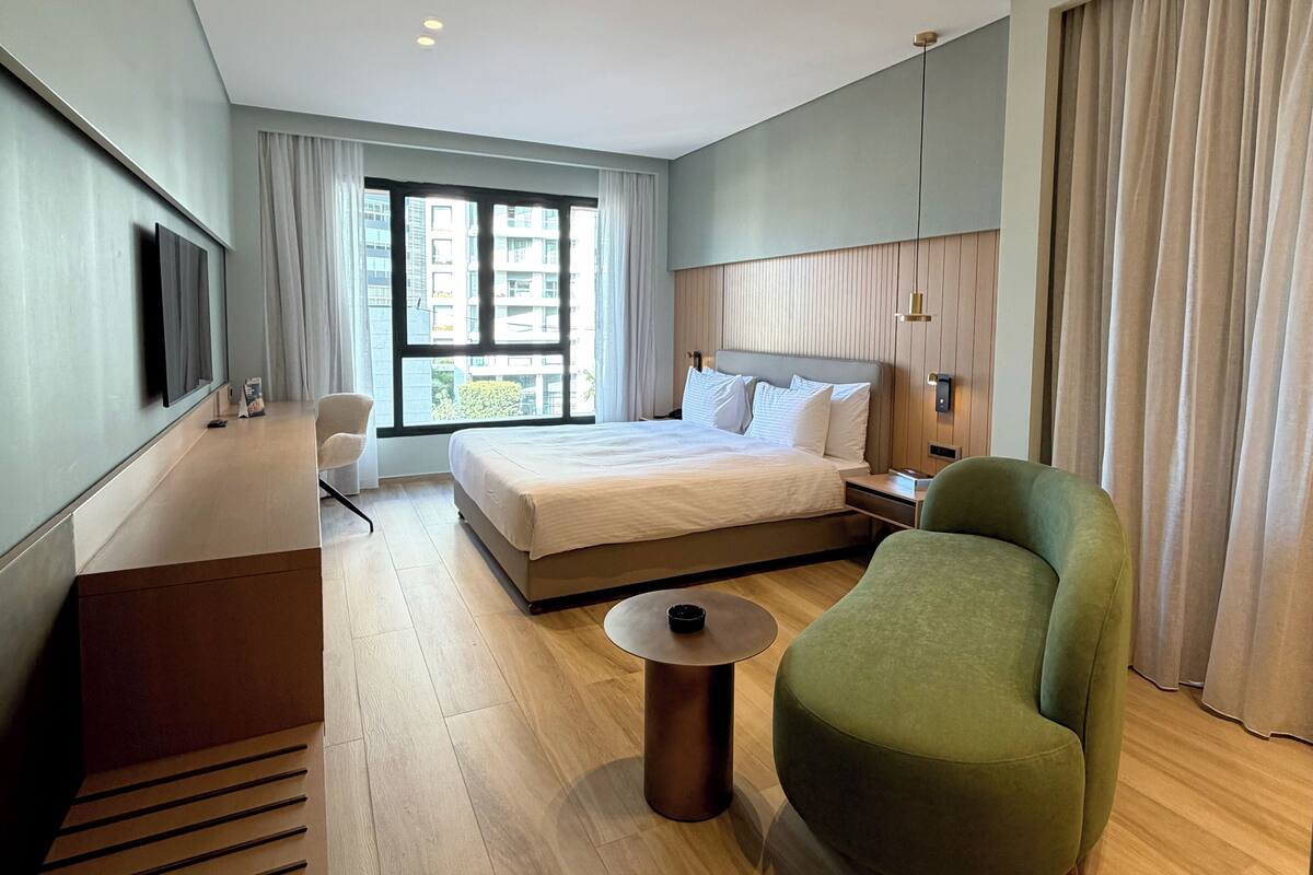 Quarto de hotel moderno com uma cama grande, uma poltrona verde, uma escrivaninha e vista da cidade através de amplas janelas.