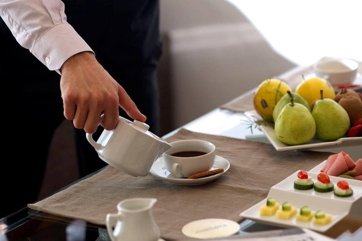 Alguém a servir café numa chávena, com um prato de fruta e pastelaria sobre a mesa.