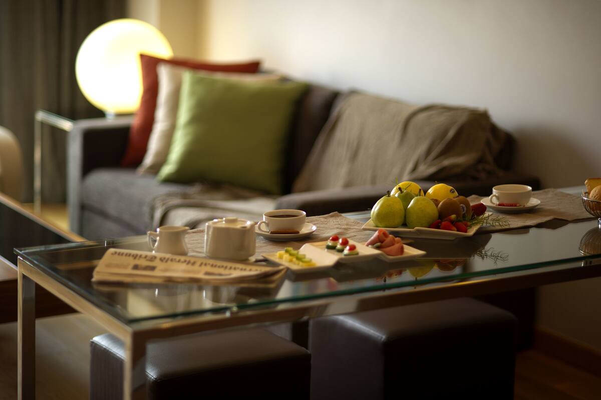 Uma sala de jantar com mesa de vidro, jornal, xícara de café e prato de frutas.