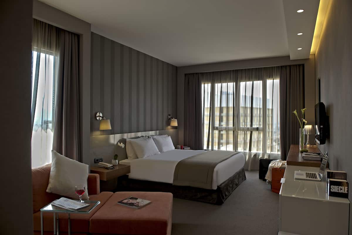 Quarto de hotel moderno com uma cama grande, uma escrivaninha com um laptop, um sofá e uma janela com cortinas.