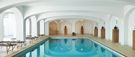 Uma grande piscina interior com vista clara para a área da piscina, rodeada por paredes e arcos brancos, e equipada com espreguiçadeiras e armários de madeira.