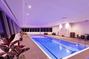 Uma grande piscina interior com piso ladrilhado, rodeada por janelas e plantas.