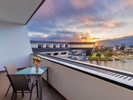 Varanda com mesa e cadeiras, vista para um corpo d'água e um complexo moderno de edifícios.