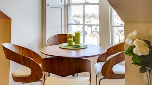 Uma área de refeições com uma mesa redonda, quatro cadeiras de madeira com almofadas brancas e um vaso de flores brancas à direita.