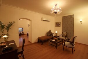Uma sala de estar com sofá, poltronas, mesa de centro, mesa de jantar e um candelabro. Há uma televisão ao fundo.