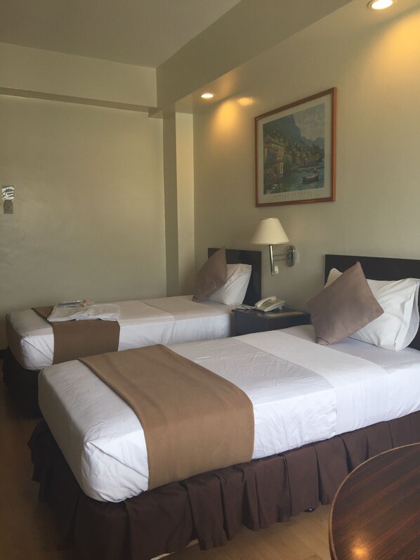 Quarto de hotel com duas camas, mesa de cabeceira com luminária, um quadro na parede e uma mesa de madeira.