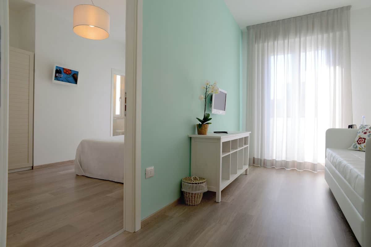 Um quarto com uma cama, uma mesa consola branca com uma planta, uma televisão montada na parede e uma janela grande com cortinas translúcidas.